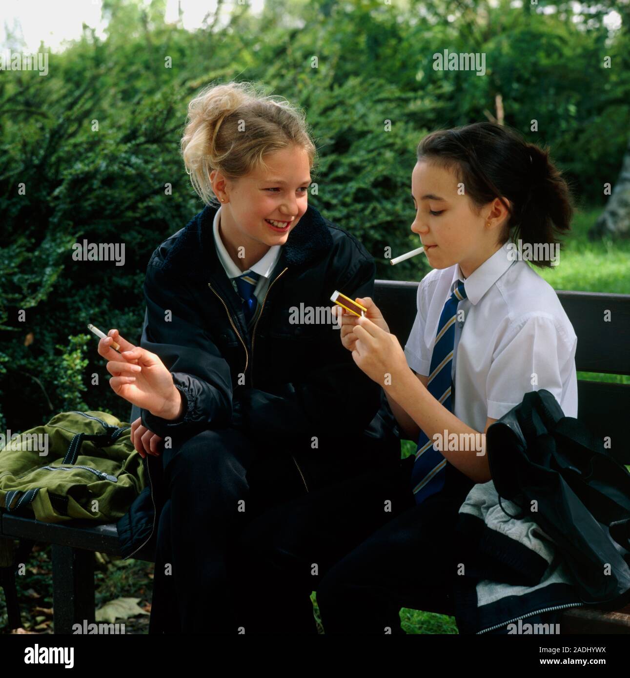 Teenage smoking. Young teenage girl lighting a cigarette with a match outdoors. Another girl is ...