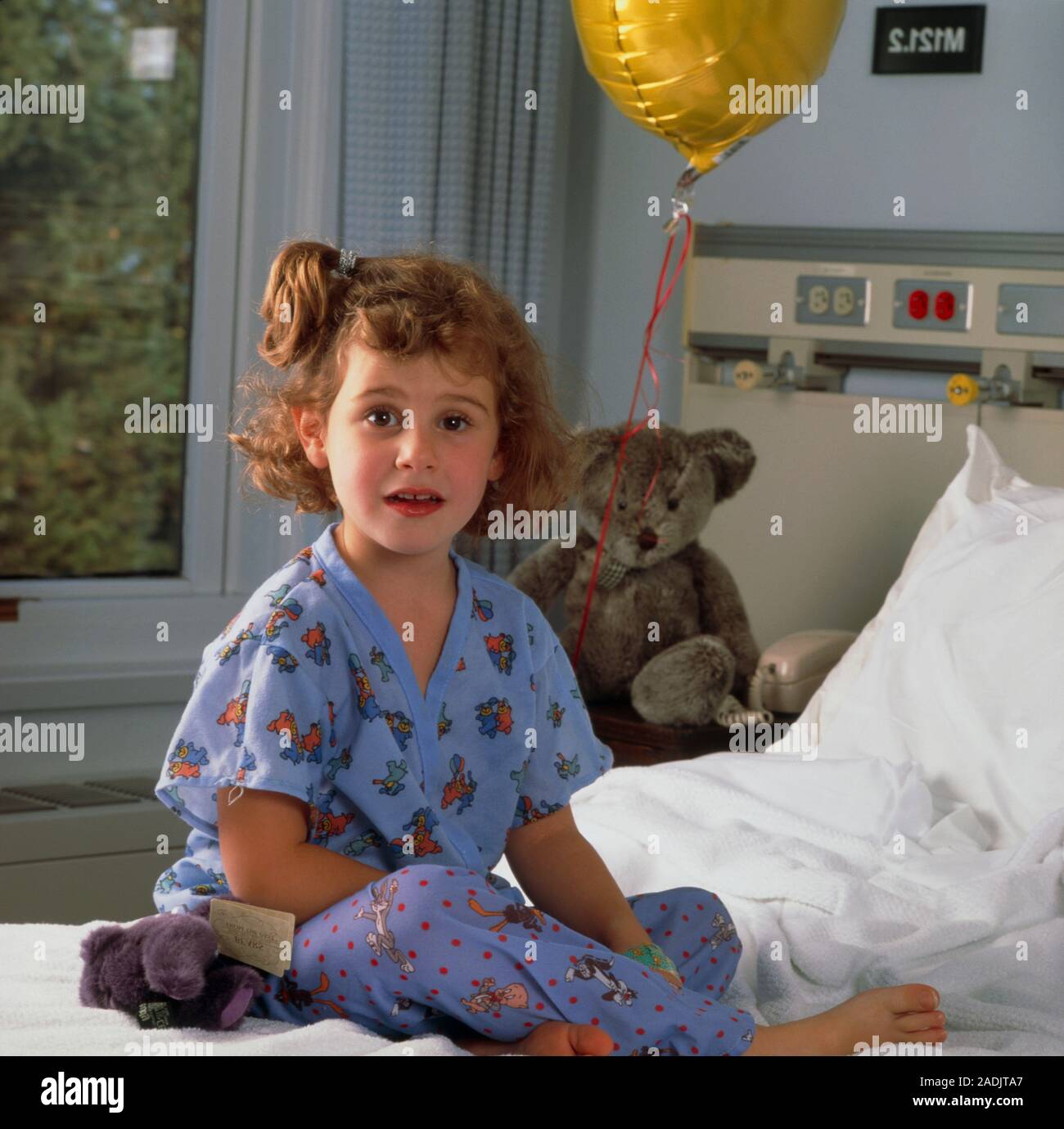 Paediatric care. Young girl sits in her pyjamas on a bed in the ward of ...
