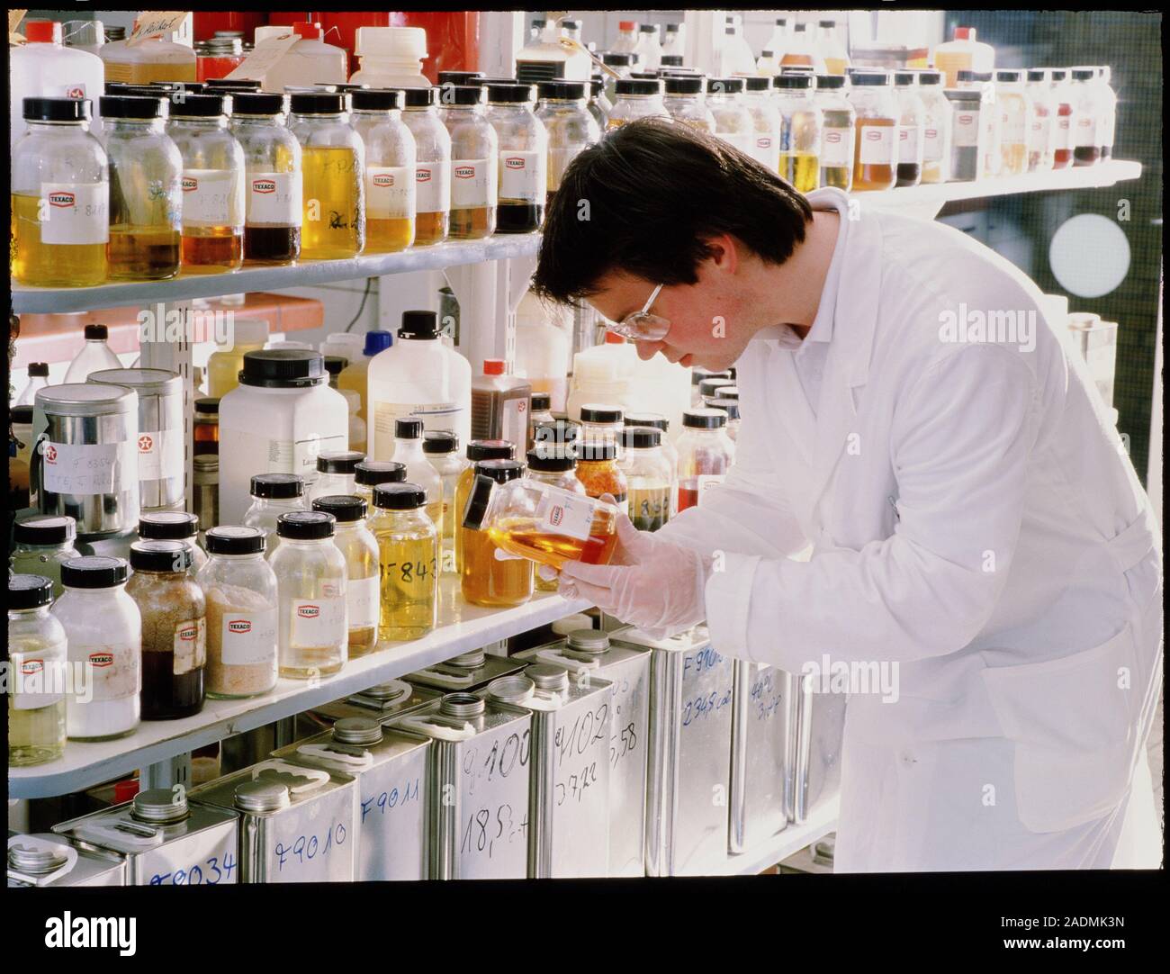 Oil analysis. Technician with shelves of mineral oil samples in a ...