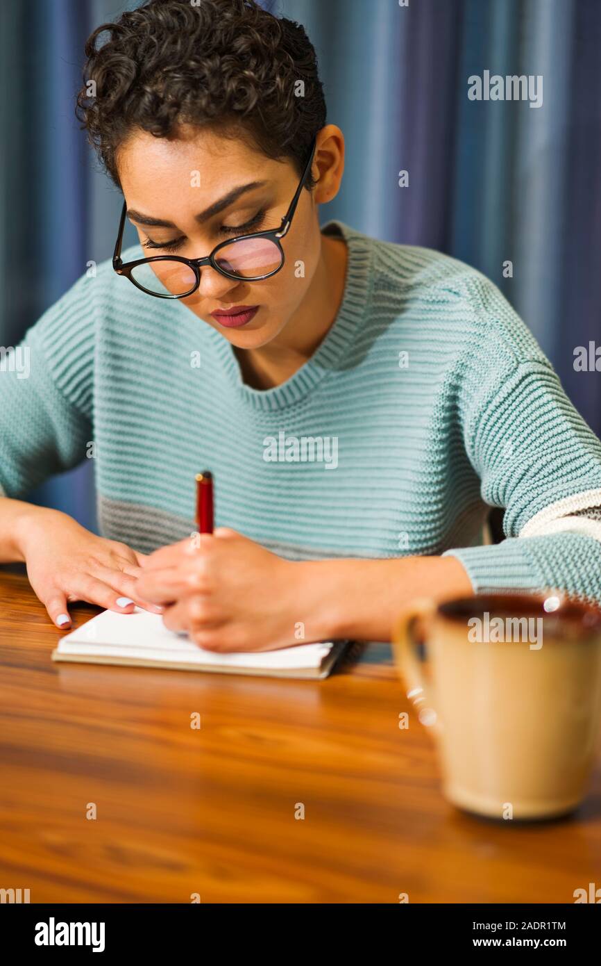 A woman wearing glasses sits at a table drinking coffee and writing in ...