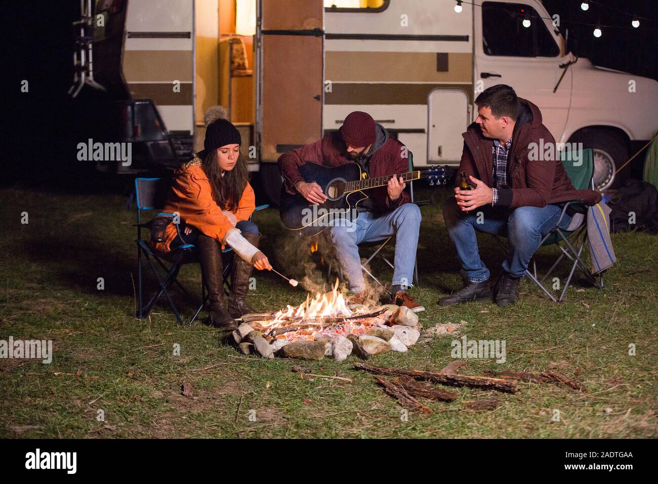 Man playing on guitar around camp fire while his friends ar roasting ...