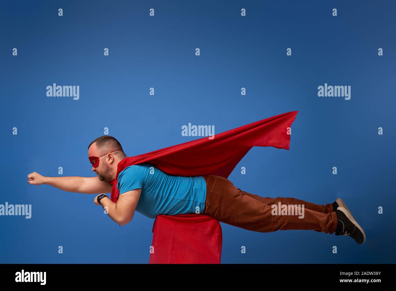 Male in red superhero cloak flying on empty blue background in studio ...