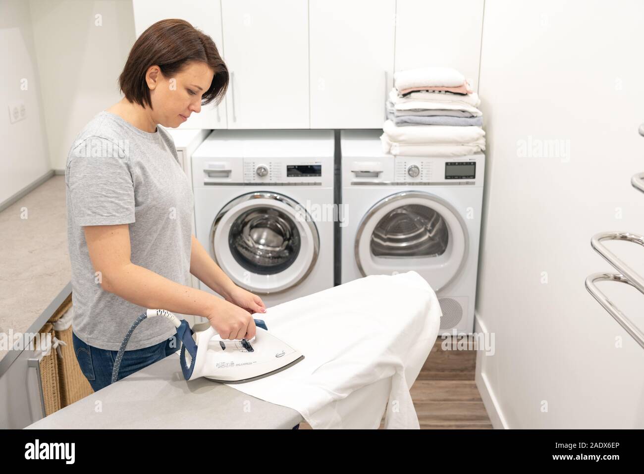 Woman ironing white shirt on board in laundry room with washing machine ...