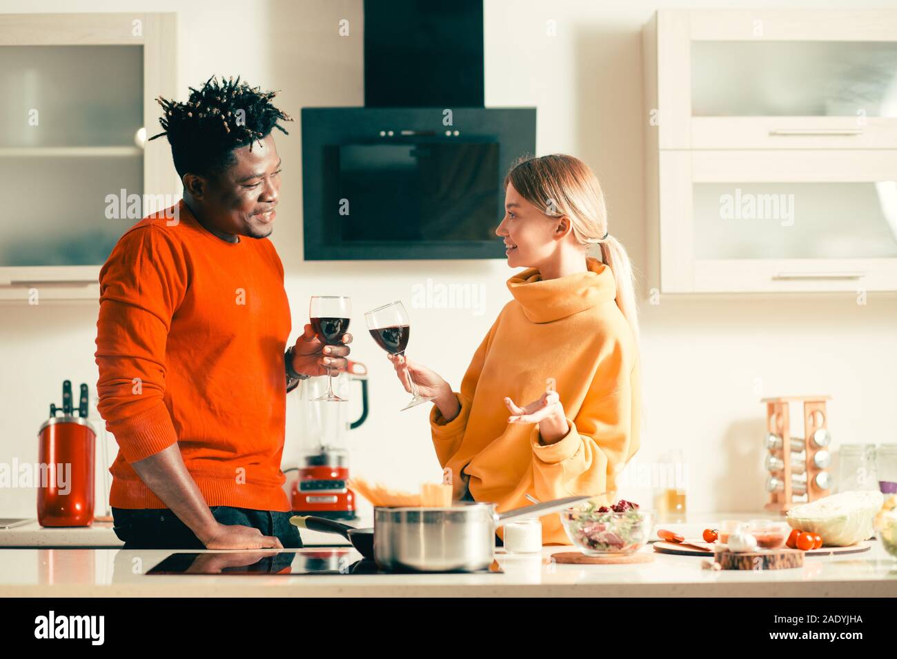 Two people talking in the kitchen while holding glasses of red wine ...