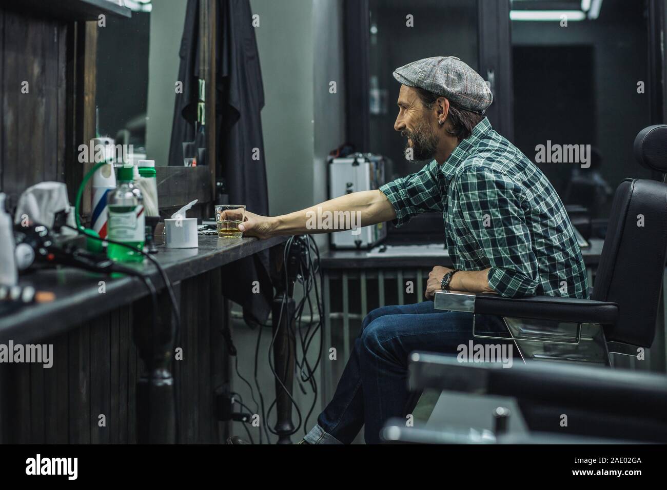 Cheerful man in flat cap drinking alcohol while being in barber shop ...