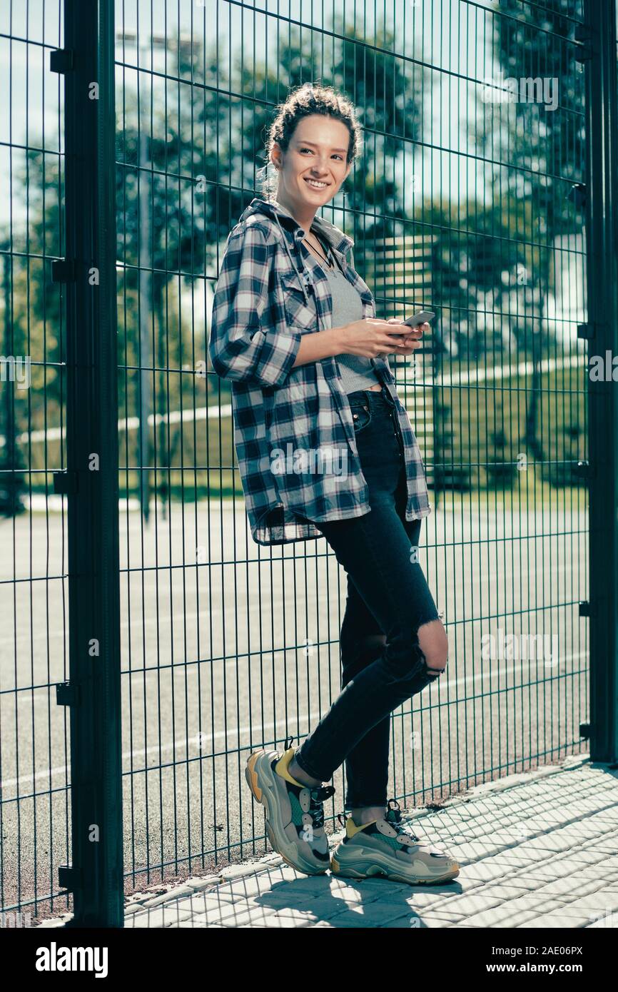 Full length of young lady leaning on the chain link fence and smiling ...