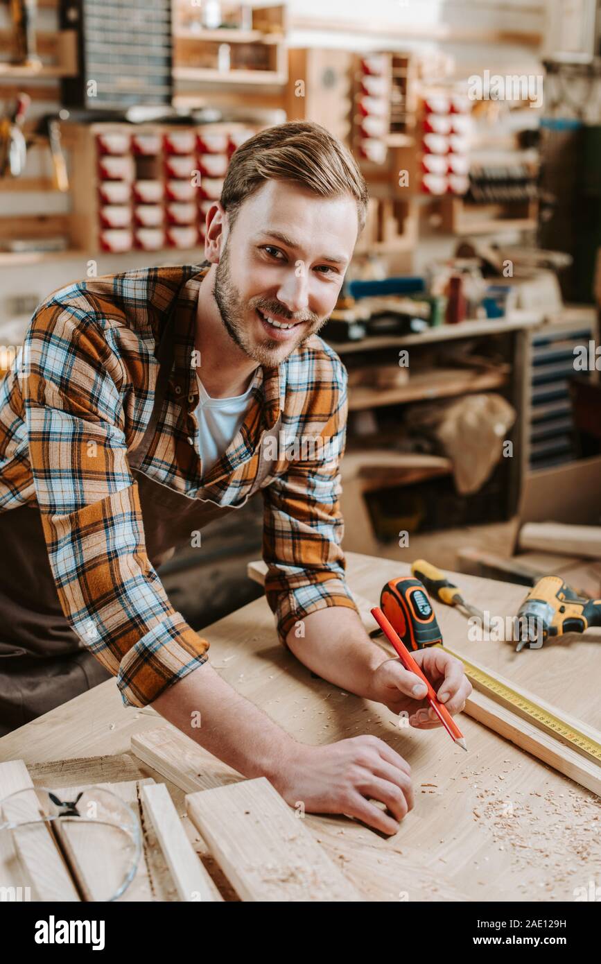 cheerful woodworker holding pencil near wooden plank and measuring tape ...