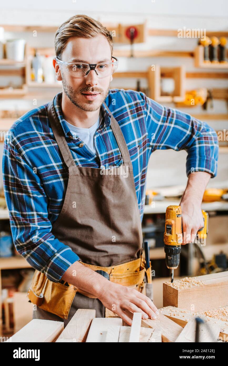 selective focus of handsome woodworker in goggles holding hammer drill ...