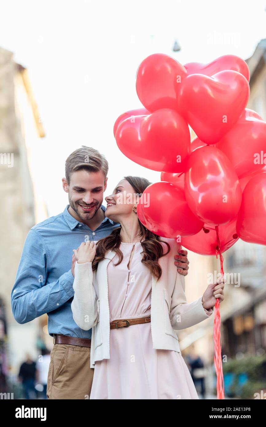 smiling man embracing happy girlfriend holding red heart-shaped ...