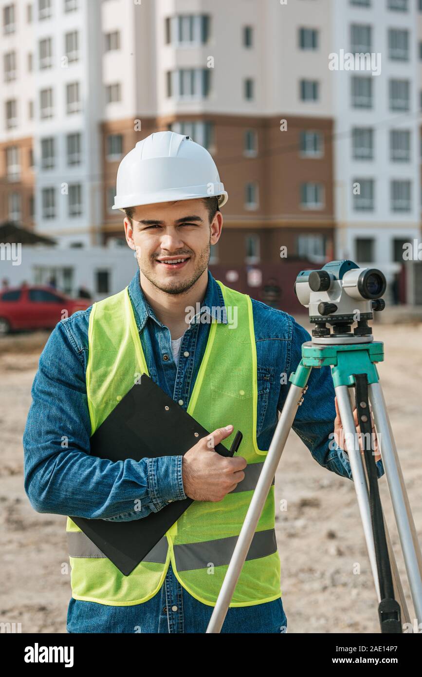 Surveyor holding clipboard and digital level and smiling at camera on ...