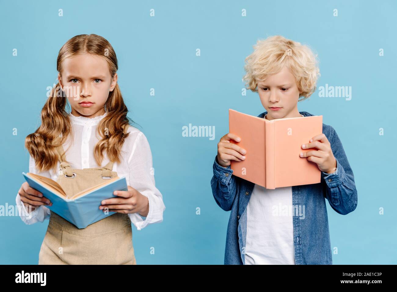 sad and cute kids looking at camera and reading book isolated on blue ...