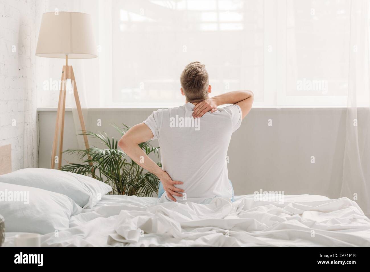 back view of man sitting on white bedding and suffering from back pain ...