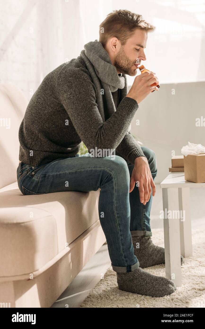 sick man in warm scarf and socks sitting on sofa and drinking cough ...