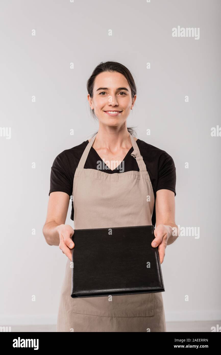 Happy young waitress of cafe in apron giving you menu in black leather ...