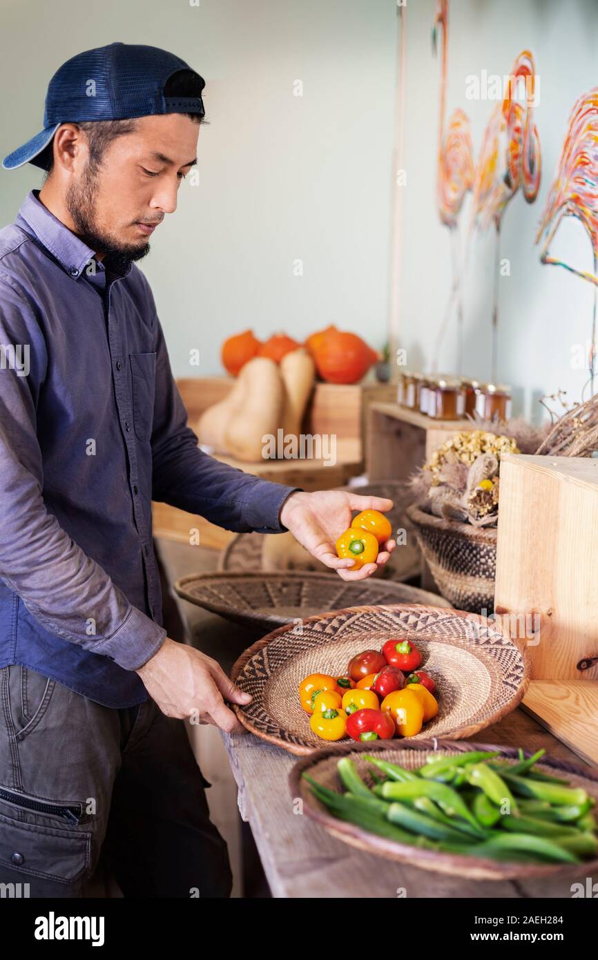 Japanese man wearing cap standing in farm shop, holding bowl with fresh ...