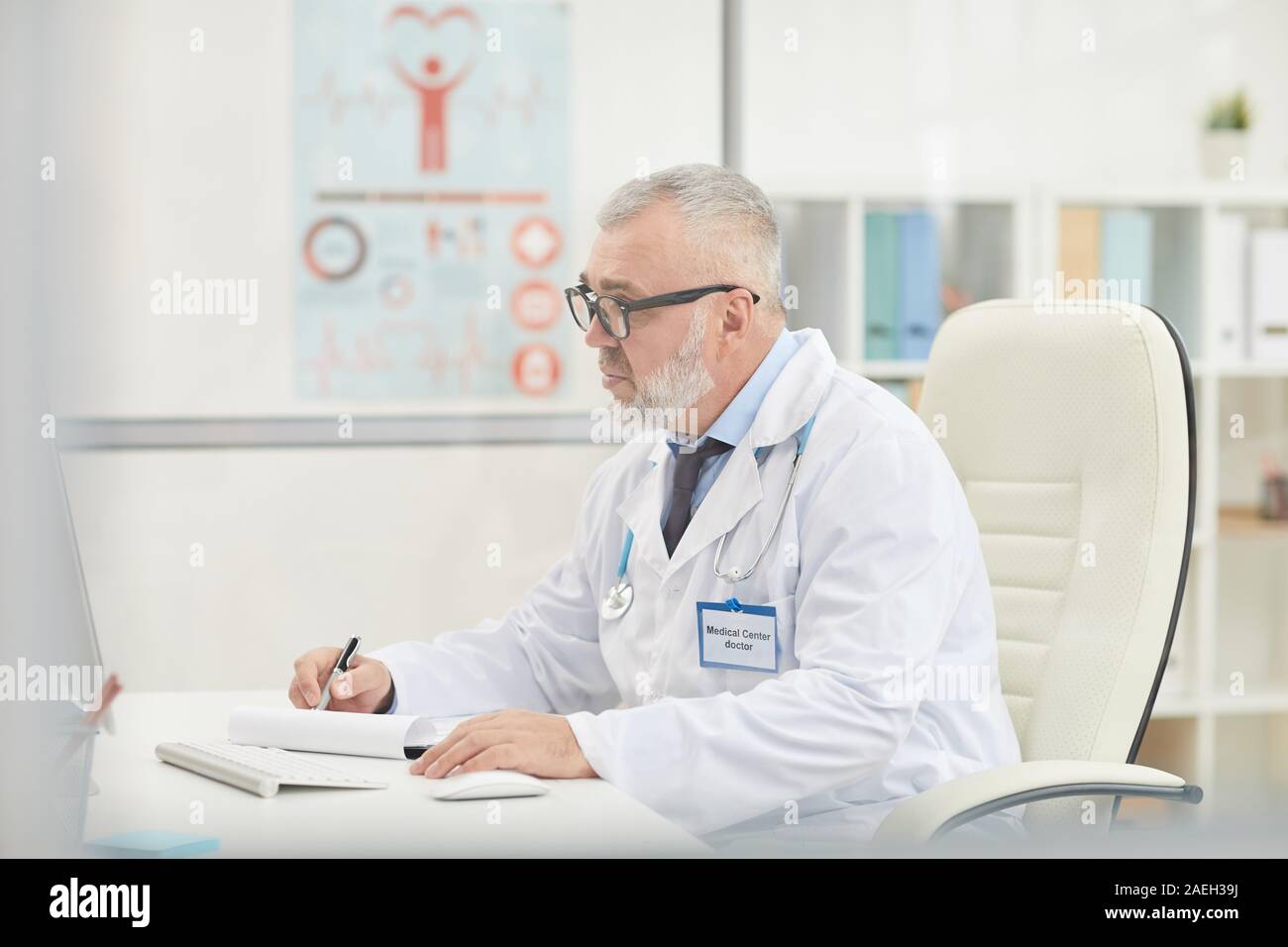 Mature doctor in eyeglasses sitting at his workplace looking at ...