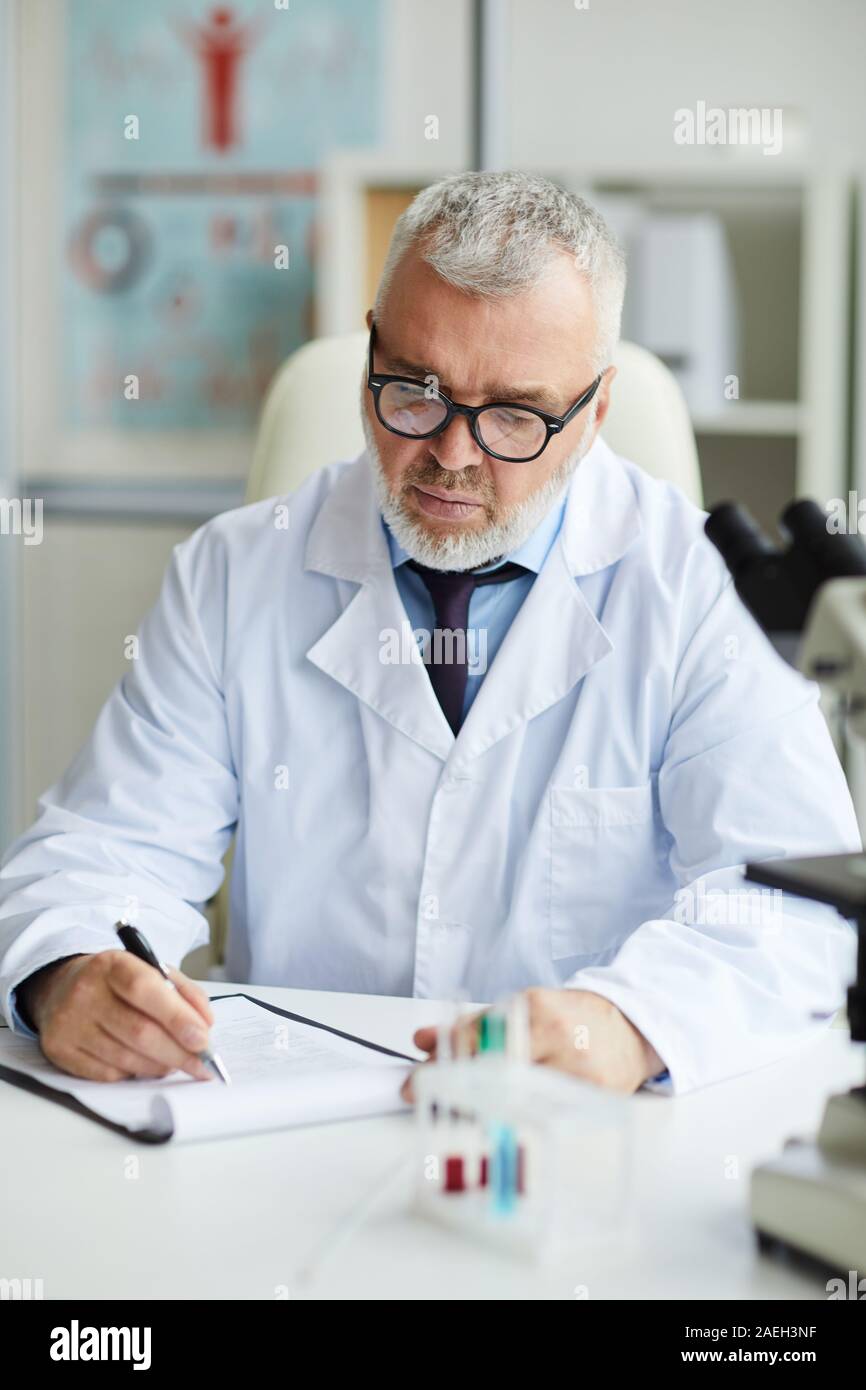 Serious mature scientist in eyeglasses sitting at the table and making ...