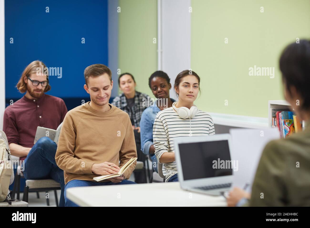 Group of young people sitting on chairs with notebooks and listening to ...