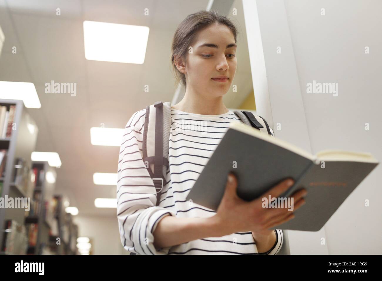 Serious student with backpack behind her back standing and reading a ...