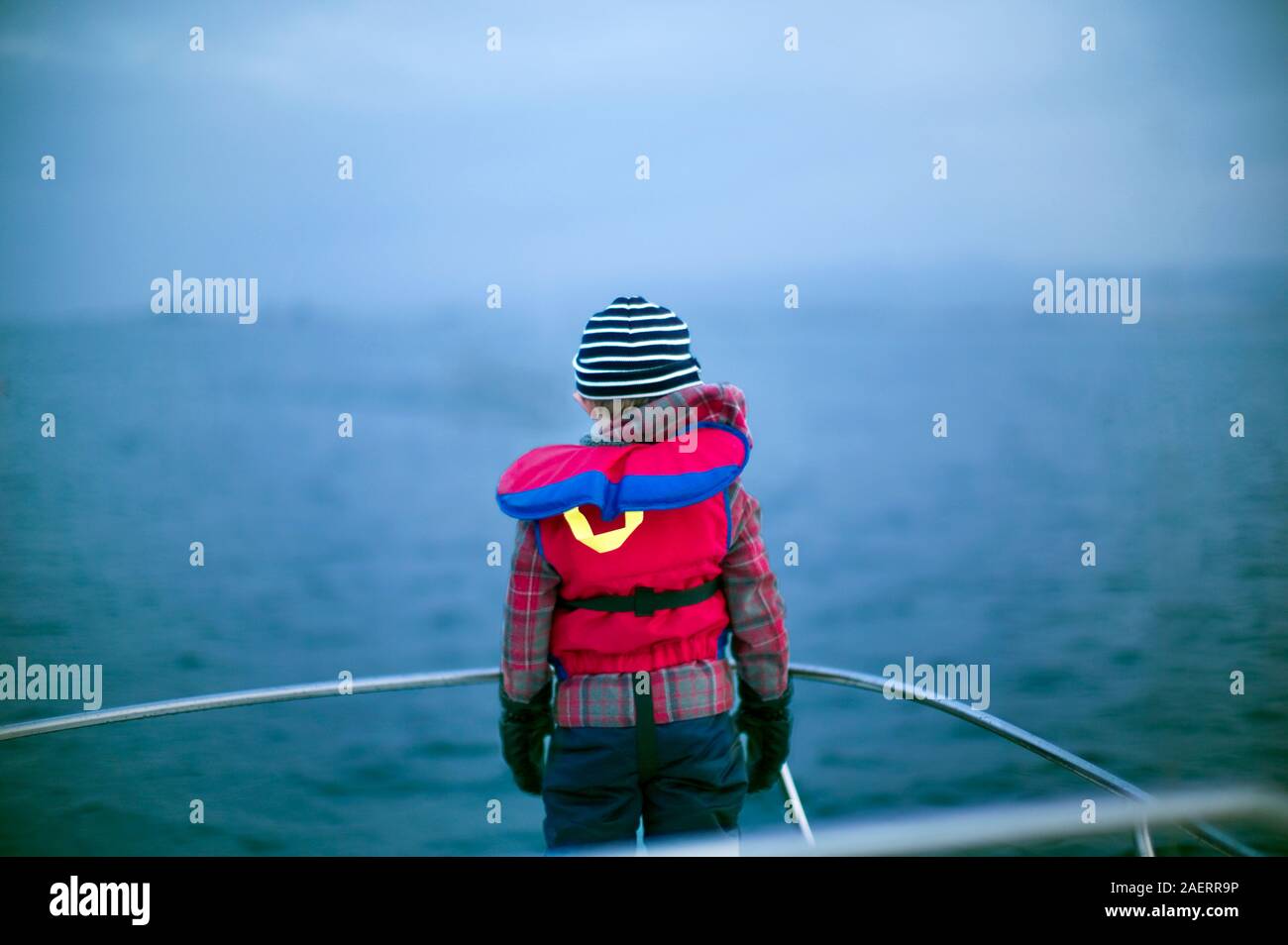 Small boy wearing a life jacket looks off the edge of boat into the sea ...