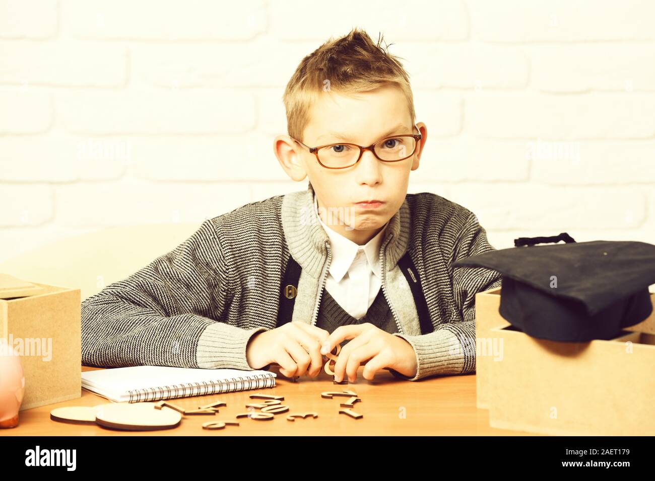 young cute pupil boy in grey sweater and glasses sitting at desk with ...