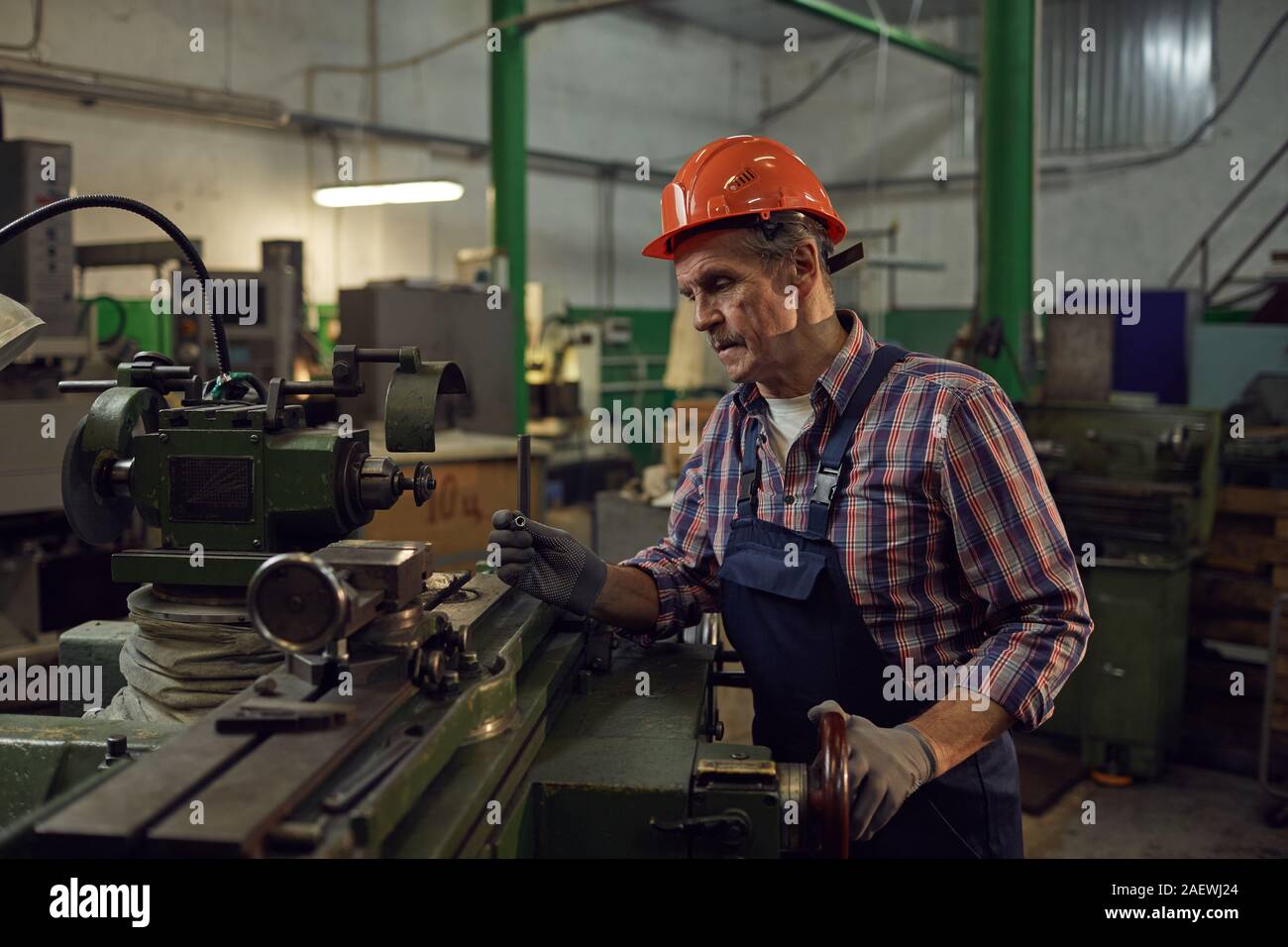 Mature man in overalls and in work helmet using lathe to do his work in ...