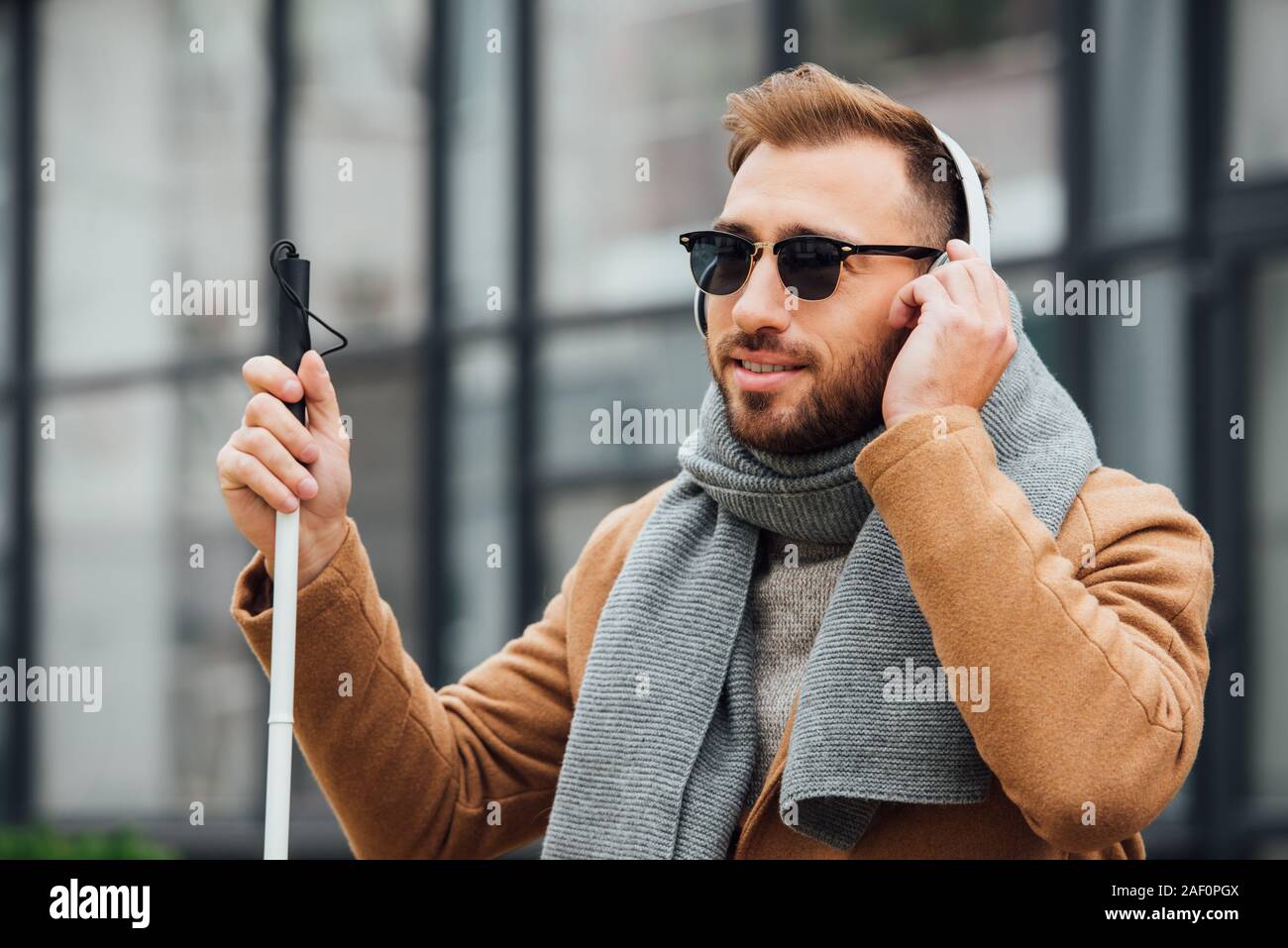 Smiling blind man using headphones and holding walking stick outdoors ...