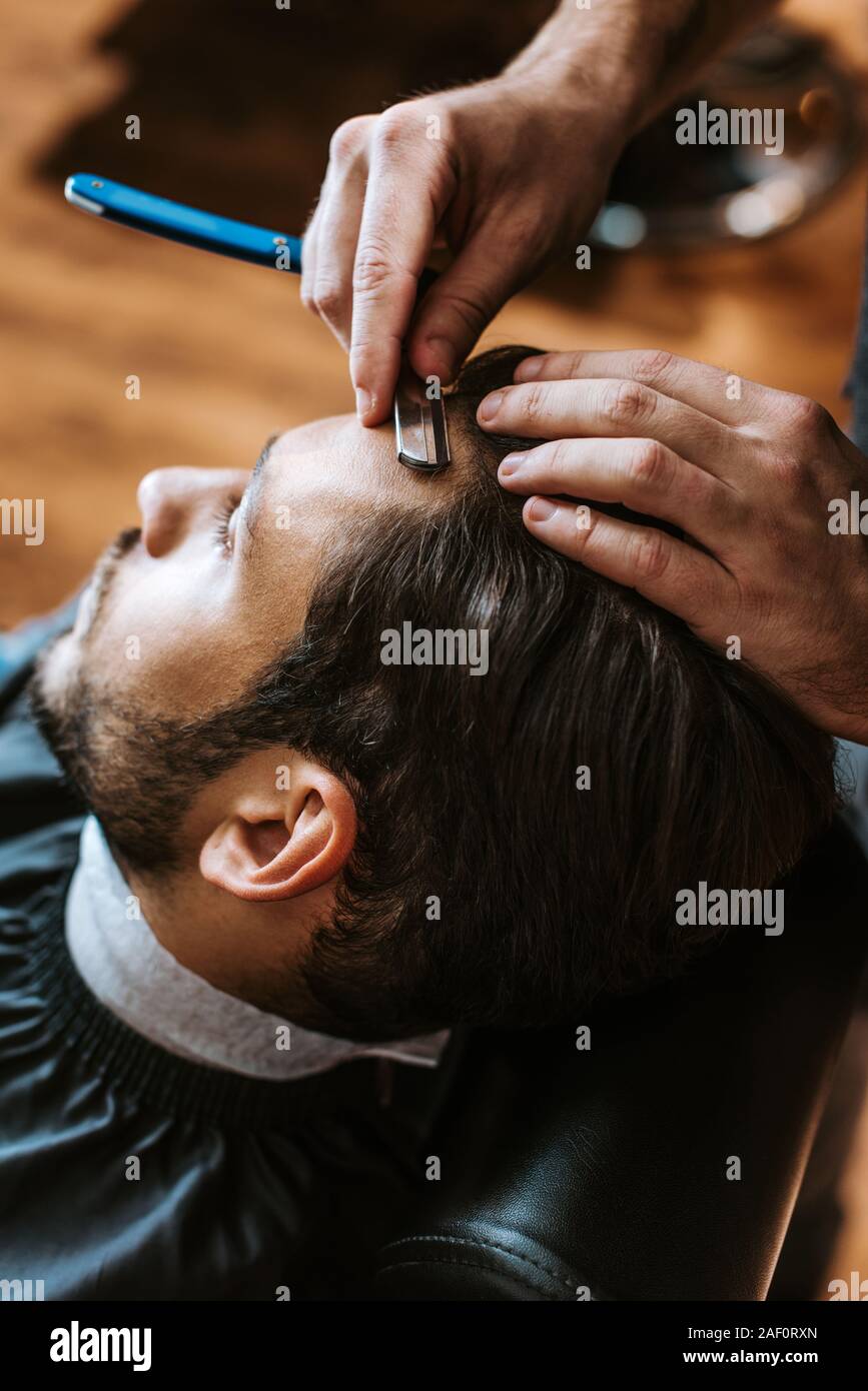 selective focus of barber holding sharp razor while doing haircut to ...