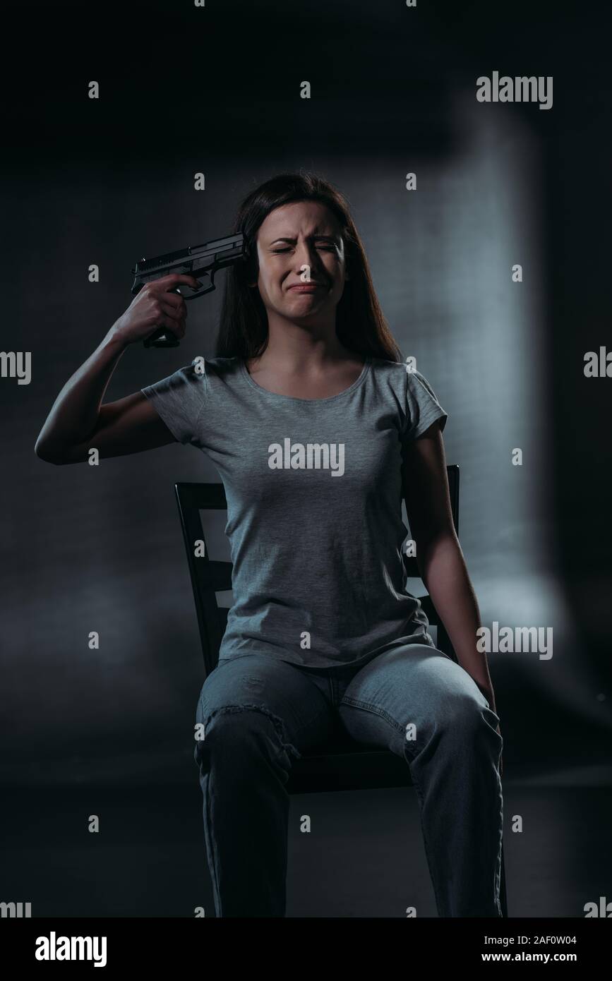 crying, despaired woman holding gun near chin while sitting on chair on ...