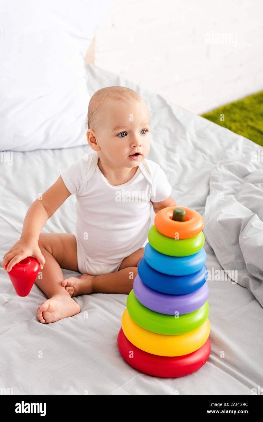 Funny barefoot child in white clothes playing with toy pyramid on bed ...