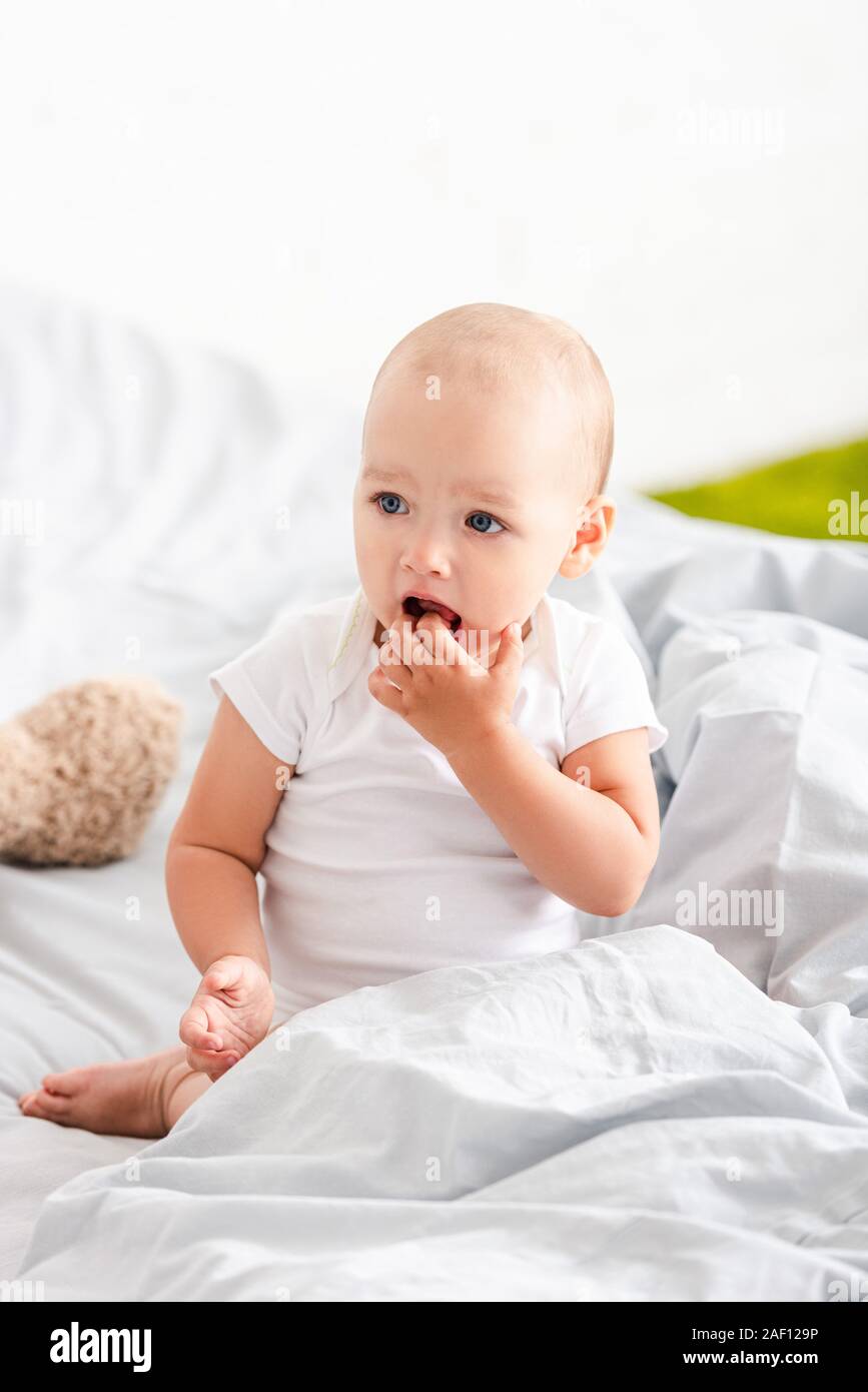 Cute barefoot child in white clothes sitting on bed and taking fingers ...