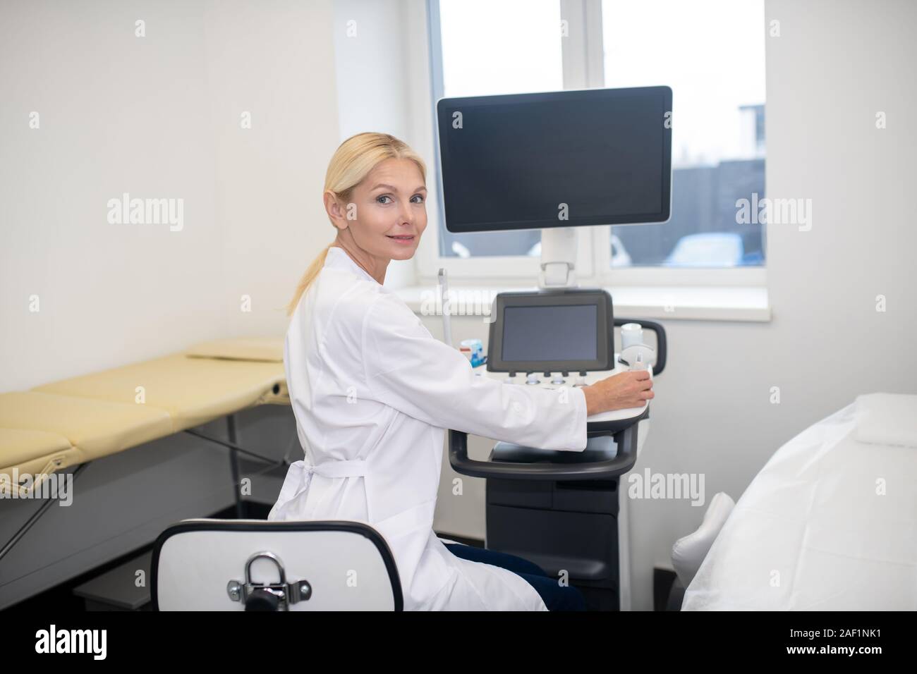 Blond pretty ultrasound specialist in white robe sitting at the ...