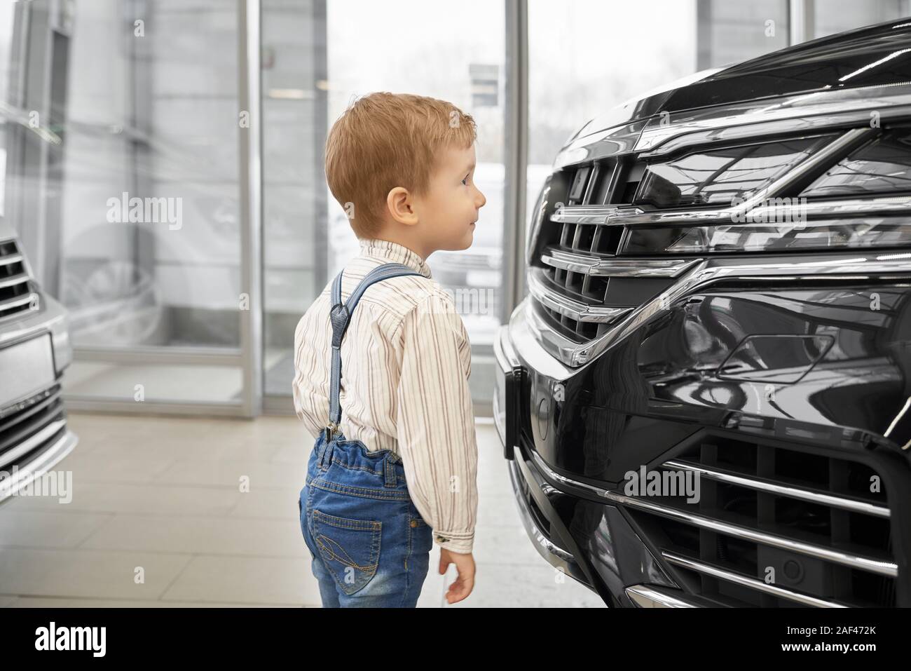 Side view of cute little boy standing near big black car in auto salon ...