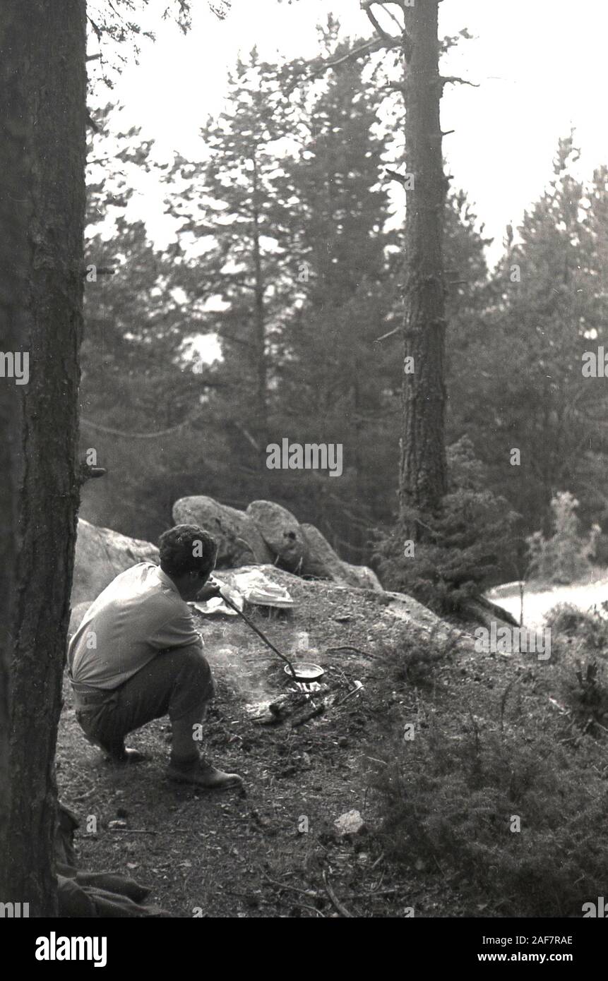 1930s, historical, a man on a rocky ledge in a forest cooking on a ...