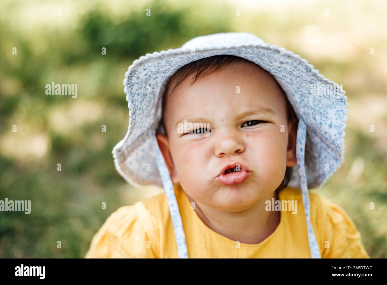 Caucasian baby girl is grimacing to the camera in hat and yellow dress ...