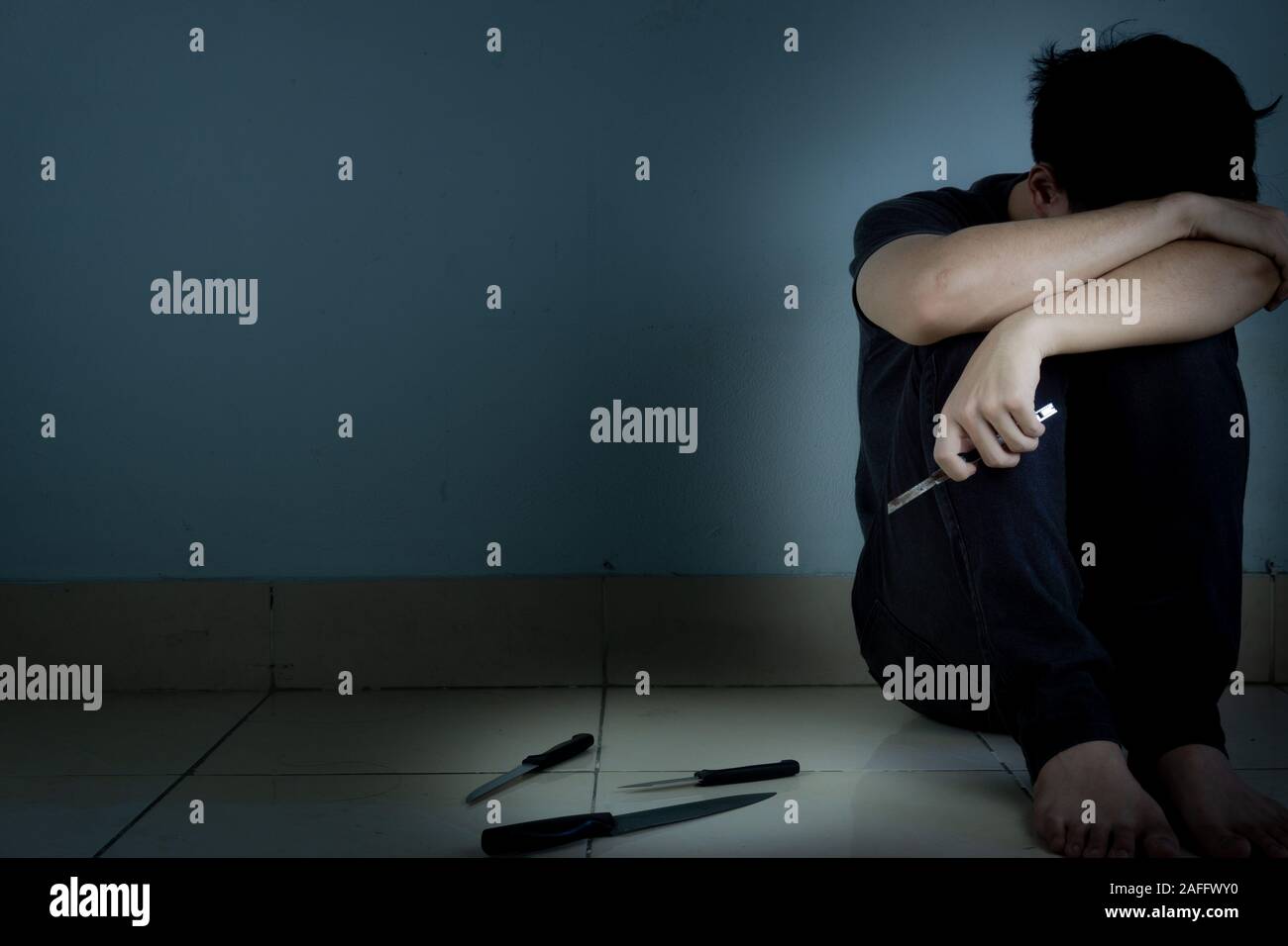 sad man hug his knee and cry sitting alone and holding a cutter knife ...