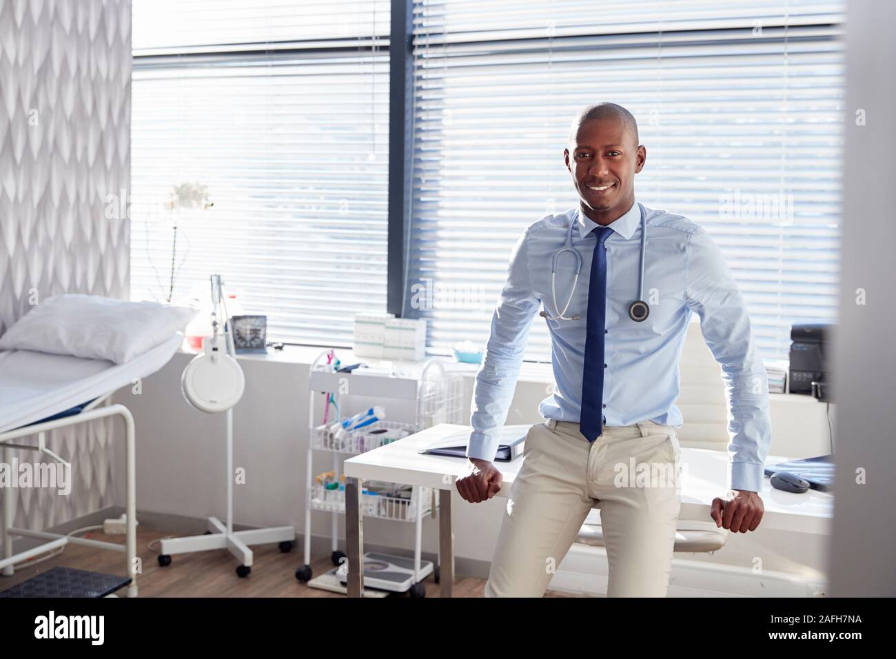 Portrait Of Smiling Male Doctor With Stethoscope Standing By Desk In ...