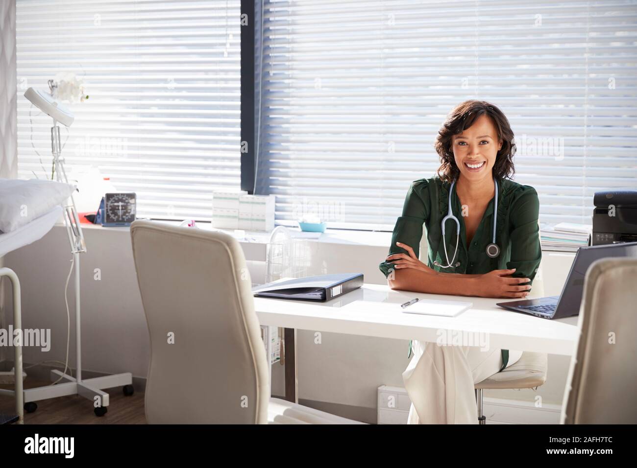 Portrait Of Smiling Female Doctor With Stethoscope Sitting Behind Desk ...