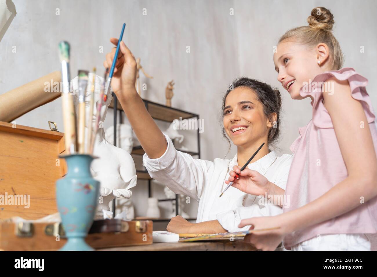 Caucasian female artist explaining little girl how to hold paintbrush ...