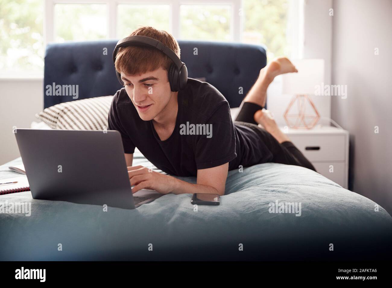 Male College Student Wearing Headphones Lies On Bed In Shared House ...