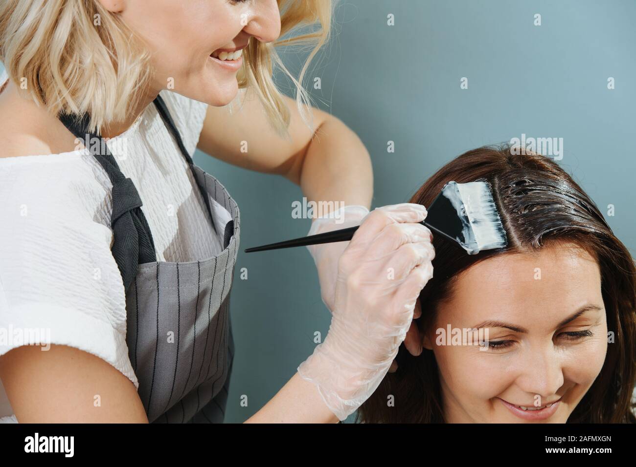 Happy hairdresser woman applying developer on client's hair over blue ...