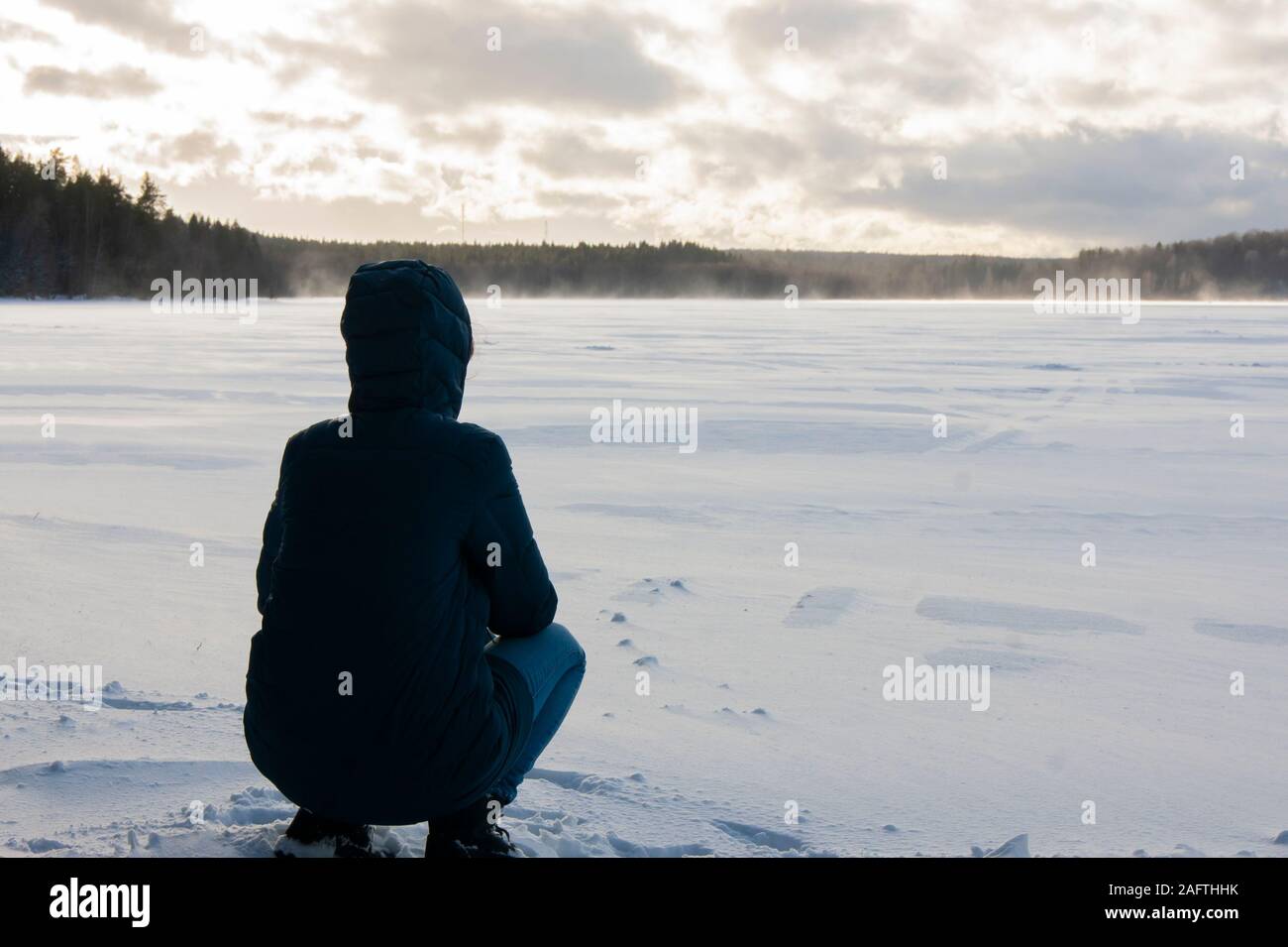 A girl sits on the shore of a winter lake. Dreaming of the warmth of ...