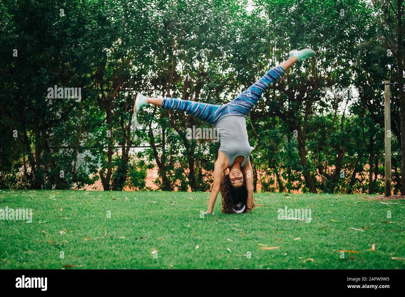 Portrait fit, carefree female personal trainer doing handstand in park ...