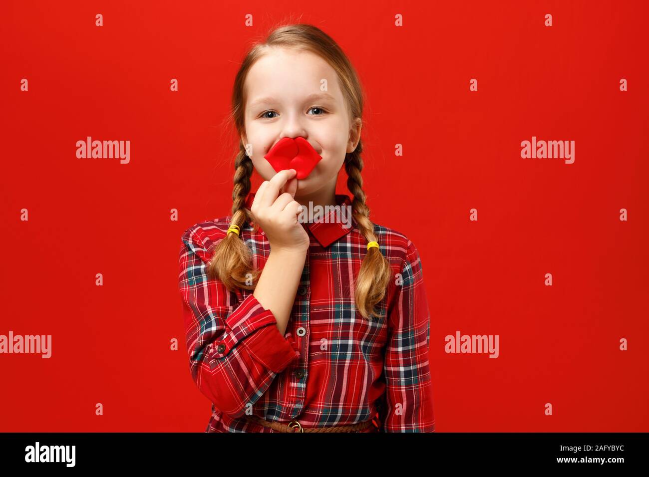 Funny cheerful little girl makes artificial fake lips on a red ...