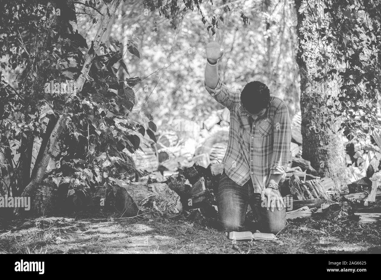 Male Down On His Knees While Reading The Bible With His Hand Up Towards The Sky In Black And
