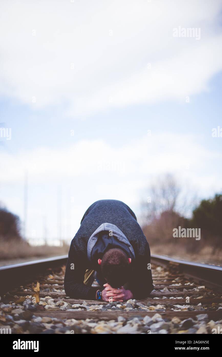Vertical shot of a male down on the ground on train track praying with ...