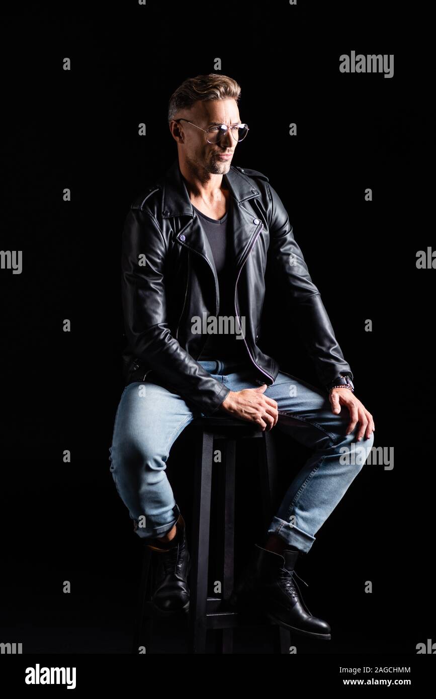 Full length of stylish man in jeans and leather jacket sitting on chair ...