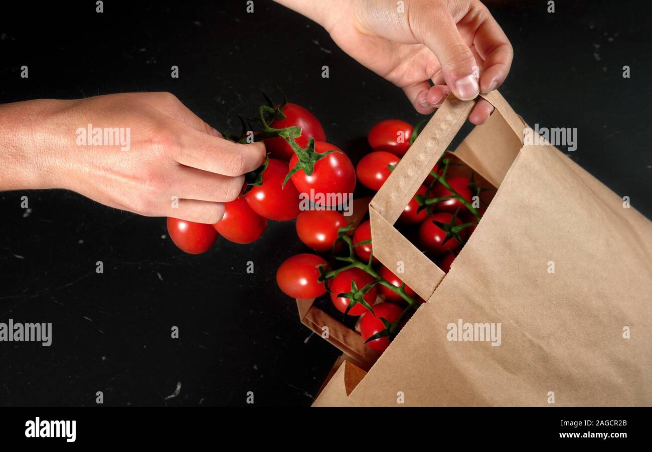 Woman hand taking fresh cherry tomatoes with green vines from brown paper shopping bag over ...