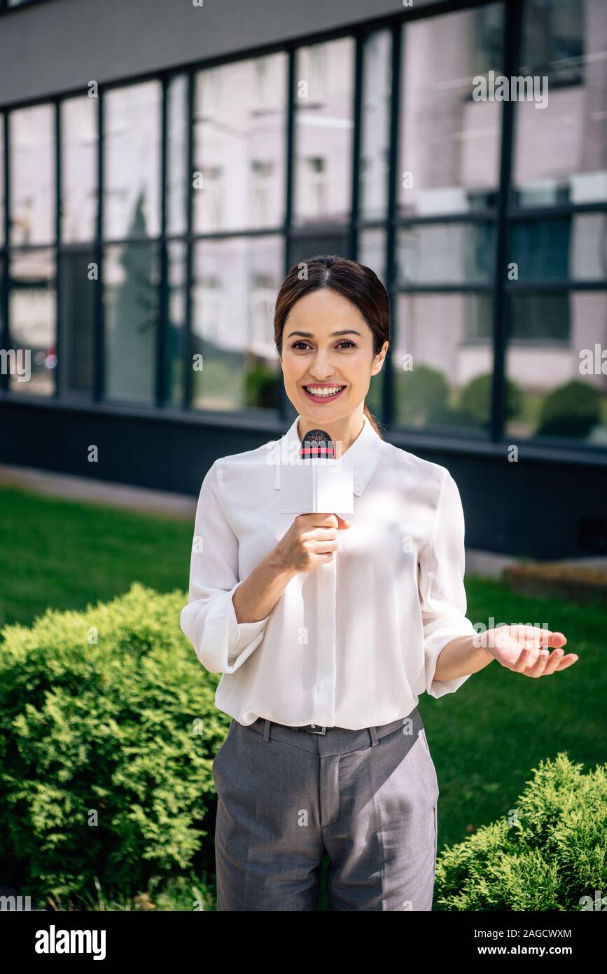 attractive journalist in formal wear holding microphone and speaking ...