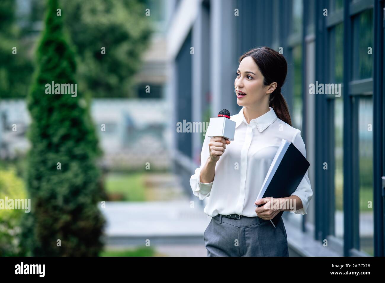 attractive journalist in formal wear holding microphone and speaking ...